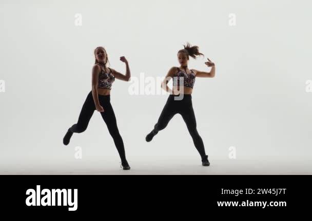 Two athletic cheerleaders in shiny tops and leggings are dancing in sync on a white studio ...