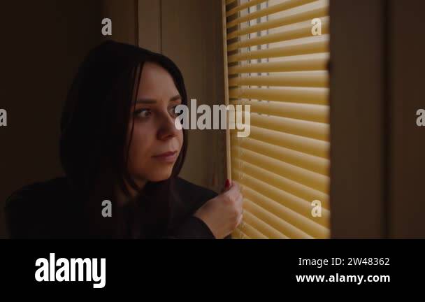 Close up of young woman watching through window blinds at home ...