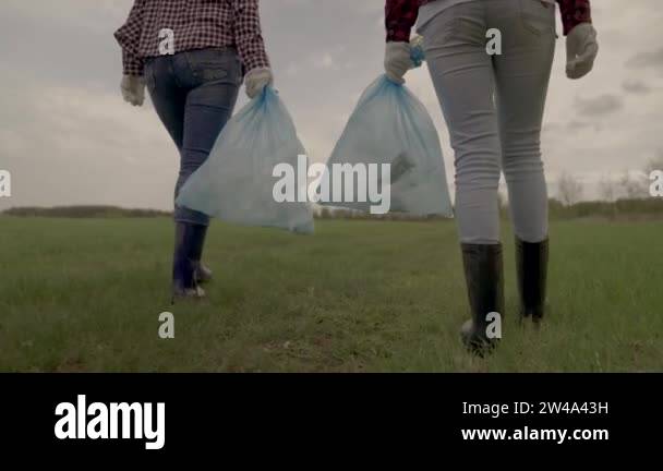 Girls walk on a green field in rubber boots and hold garbage bags in ...
