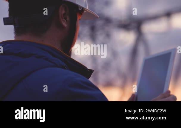 An electrician with a tablet works by a high-voltage tower at sunset in ...