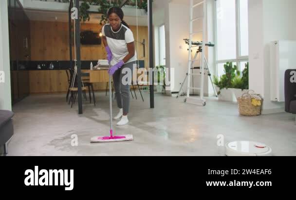 Beautiful african american woman mopping floor in living room with mop. Pretty housewife doing ...