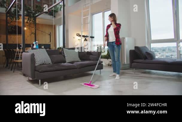 Beautiful woman in glasses mopping floor in living room with mop. Pretty housewife doing ...