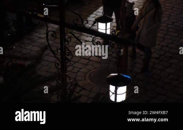 People walk along the pavement between lighting street lanterns along ...