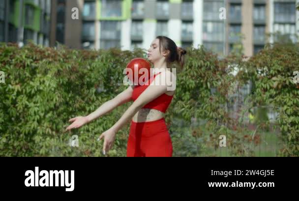 Woman in red sports costume performs callisthenics exercises with gymnastic ball outdoors ...