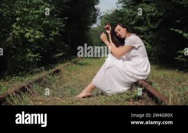 A Spectacular Young Woman In A White Dress Sits Barefoot On The Rusty Rails Of An Old Railway In