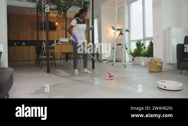 Joyful african american woman mopping floor in living room with mop. Pretty housewife doing ...
