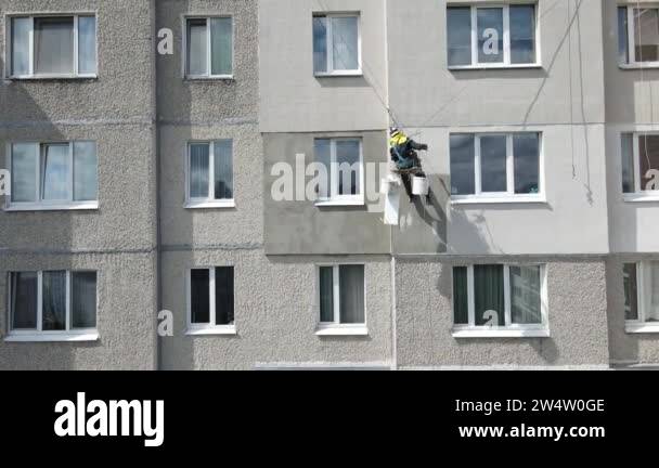 An industrial climber suspended on ropes and paints the wall of a ...
