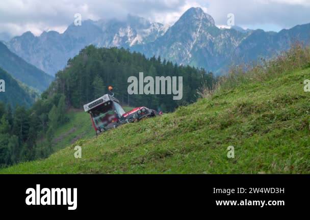 A tractor is driving down a steep hill and is cutting grass with ...