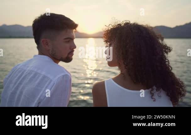 Close up of a multiracial couple kissing in backlight with the sun setting in the lake with ...
