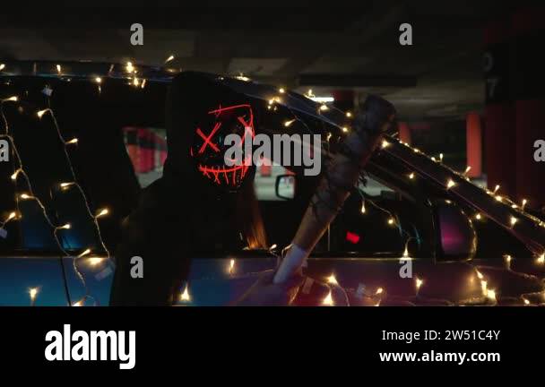 A man in a neon mask looks out of a car window in a parking lot at ...