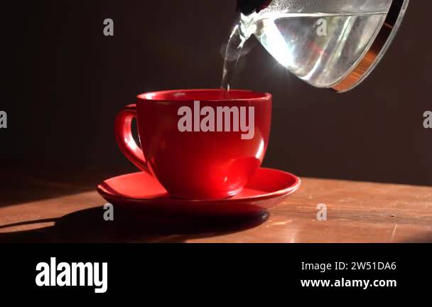Pour hot water from a clear glass teapot into a red ceramic cup on a ...