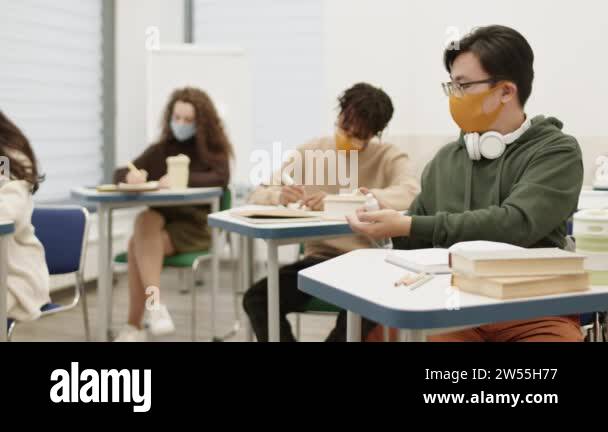 Asian Guy Wearing Glasses And Cloth Mask Sitting By Desk In Classroom Spraying Sanitizing