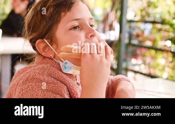 Little girl in a mask eats toast with ham and cheese in a cafe on the ...