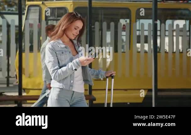 A young woman is standing at a public transport stop. She is holding a ...