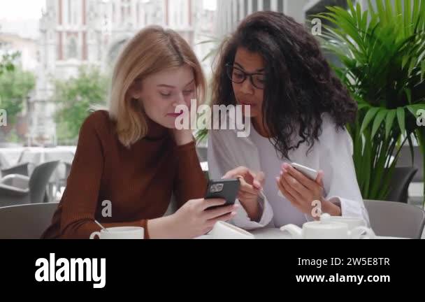 Two young beautiful women friends sitting in cafe outdoors, looking at ...