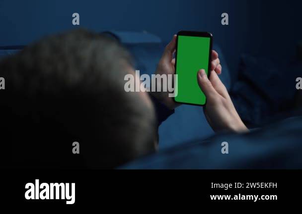 Man lying on couch using smart phone with chroma key green screen at night, scrolling through ...