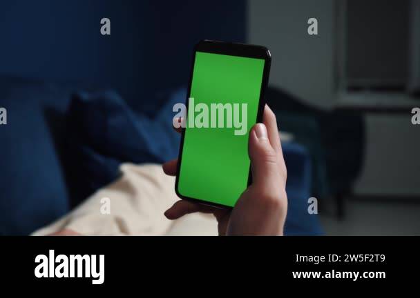 Man lying on couch using smart phone with chroma key green screen at night, scrolling through ...