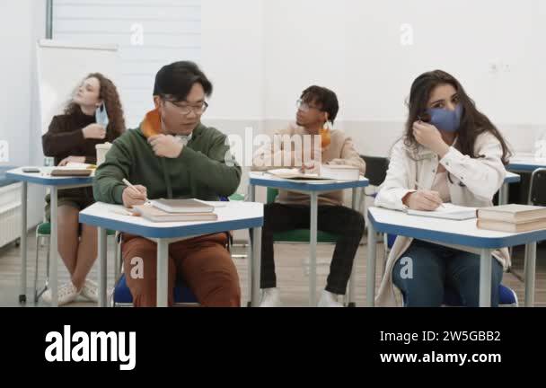 Four multiethnic students sitting by desks in classroom, taking off ...