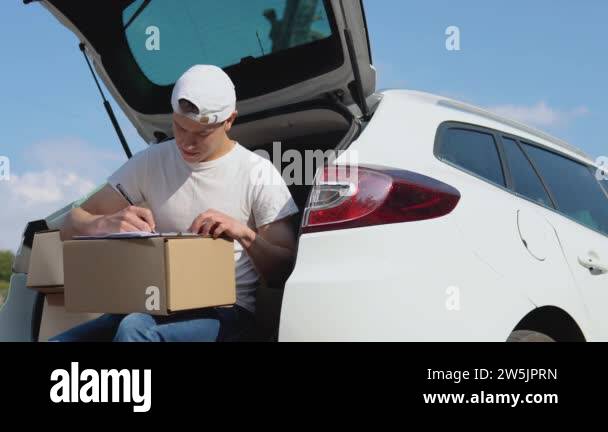 The courier sits on the edge of the open trunk, filled with boxes of ...
