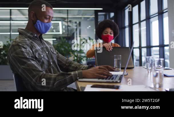 Diverse business people wearing face masks sitting using laptops going ...