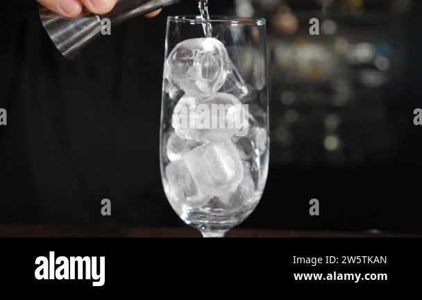 Bartender making cocktail pouring transparent alcohol liquid into glass with ice cubes. Slow ...