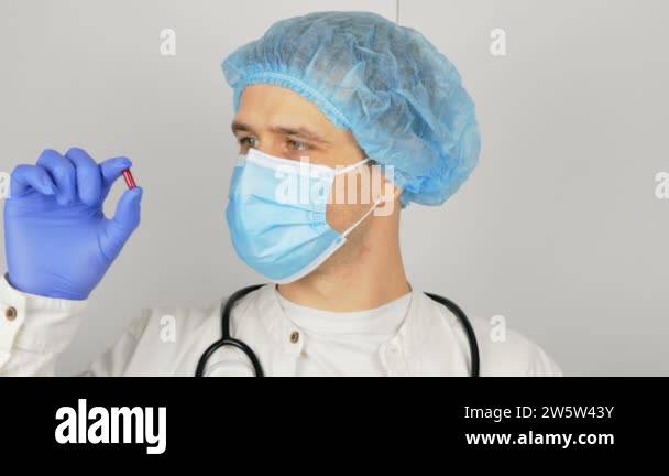 A young handsome doctor in a medical mask holds a red pill in his hands ...