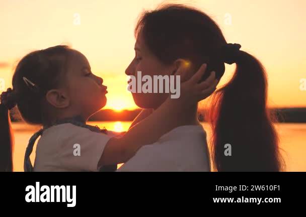 A little girl at sunset kisses a happy mother holding her in her arms ...