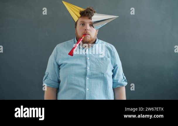 Portrait of tired young man wearing party hats blowing horn looking at ...