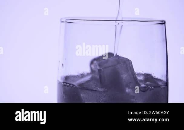 Close-up video of a glass of ice pouring into a glass of carbonated water against a glowing wall ...