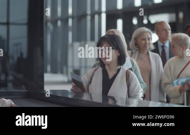 Waist-up shot of smiling young Chinese woman in glasses standing in queue at check-in counter at ...
