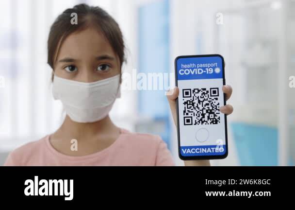 Portrait of little girl in medical mask showing smartphone in her hand with mobile application ...