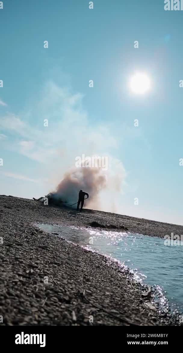 A person burns garbage in a large bonfire on the ocean, correcting the ...