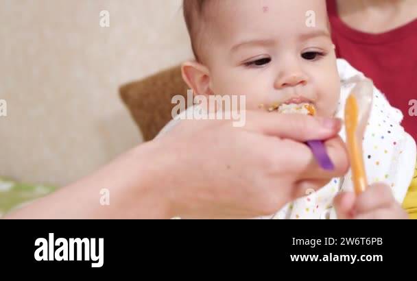 mother holds the baby in her arms and feeds her boy with a spoon ...