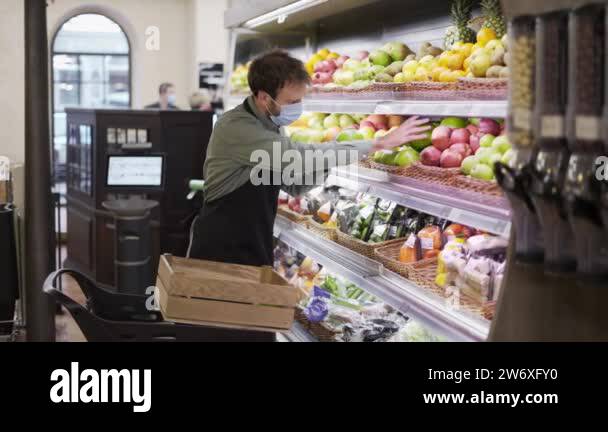 Busy salesman in a mask is putting apples on shelves at fruits section ...