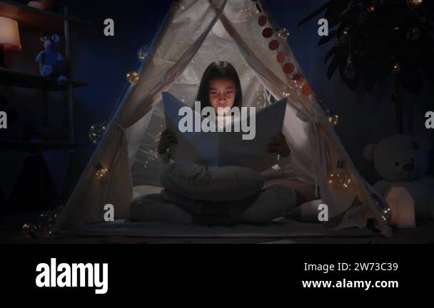 Excited girl with long black hair in cute makeshift hut opening book at ...