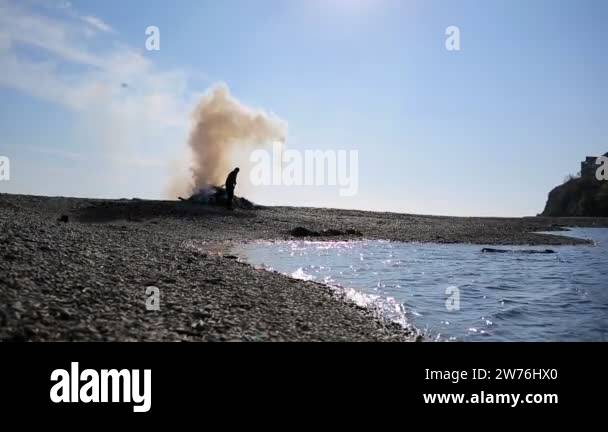 A person burns garbage in a large bonfire on the ocean, correcting the ...