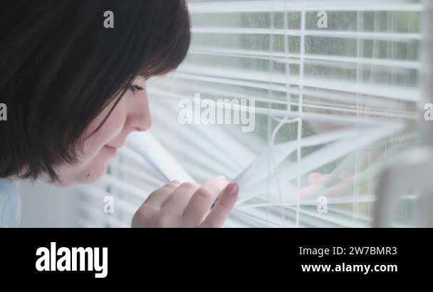 Woman looks out window through the blinds. Girl hiding in house behind ...