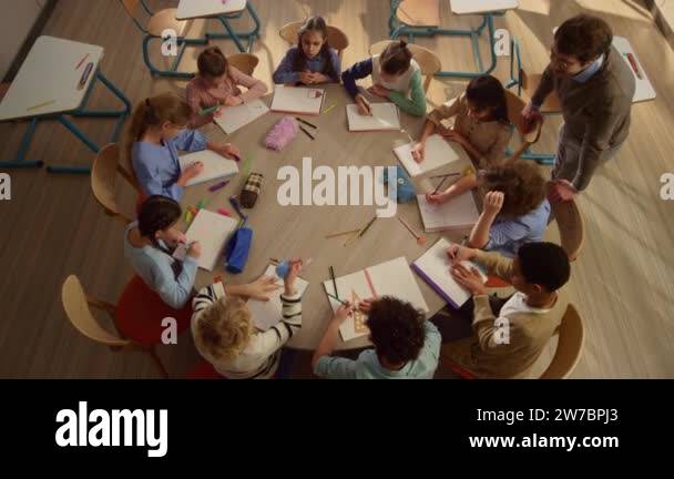Schoolchildren having lesson at round table. Male teacher helping kids ...