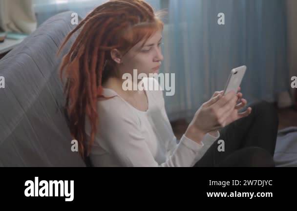 A red haired girl with dreadlocks is sitting on the couch at home ...