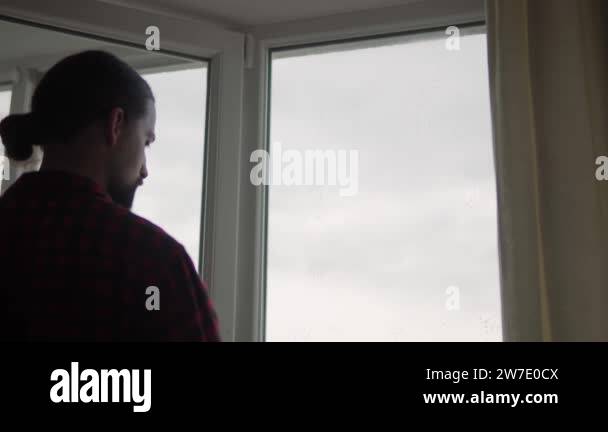 Washing window glass at home. A young long-haired man with a beard is ...