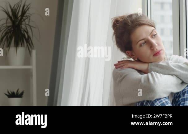 Young thoughtful woman sitting on sill, hugging her knees and looking ...