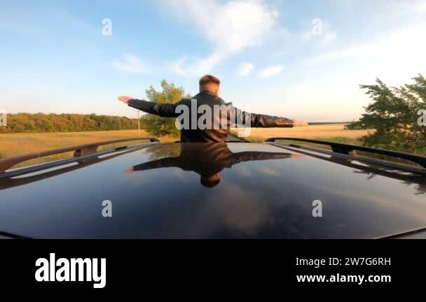 Young guy standing out of car sunroof with hands to sides and flying ...