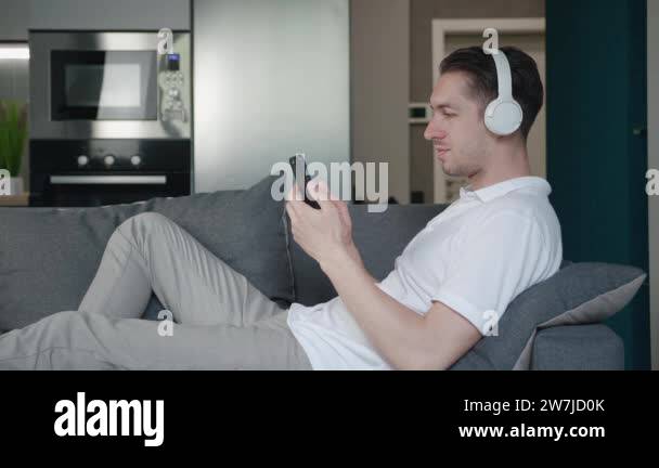 Portrait of cheerful young man lying on the sofa and using smartphone ...