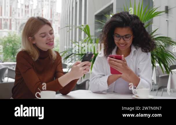 Two young beautiful women friends sitting in cafe outdoors, looking at ...