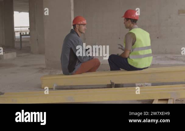 Rear view of two Caucasian builders wearing orange helmets, sitting on beam in building under ...