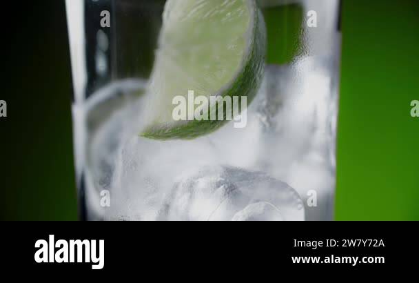 Slice of lime is added to the highball glass with cocktail and ice cubes on the green background ...
