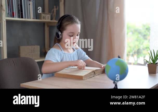 a girl studying at a remote online school listens to a lesson in headphones, reading a book with ...