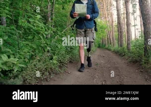 Disabled Tourist With Prosthetic Leg Walks Along Pedestrian Path In Forest And Uses Map For 