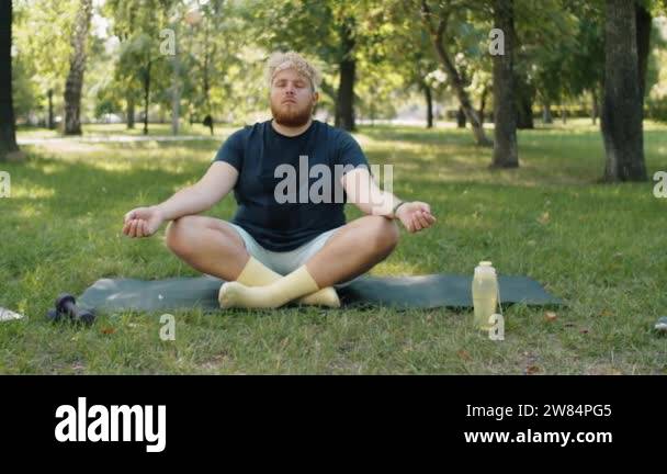 Young overweight man sitting in lotus pose with eyes closed and hands ...