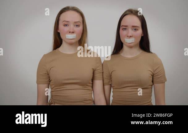 Portrait of twin sisters with taped mouth looking at camera with sad facial expression ...